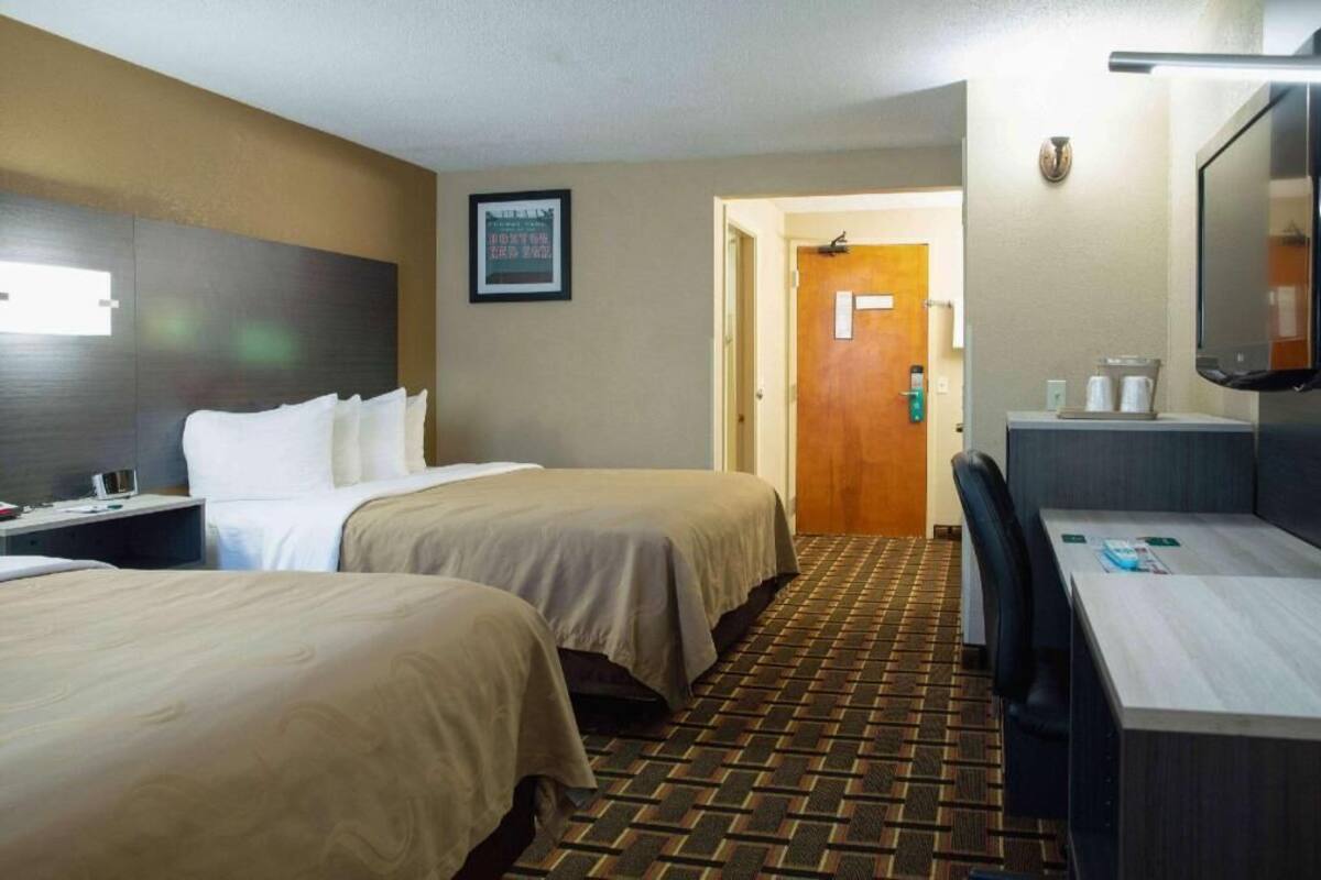 Quarto de hotel com duas camas, uma escrivaninha, uma cadeira, uma televisão e uma porta que dá para outro cômodo.