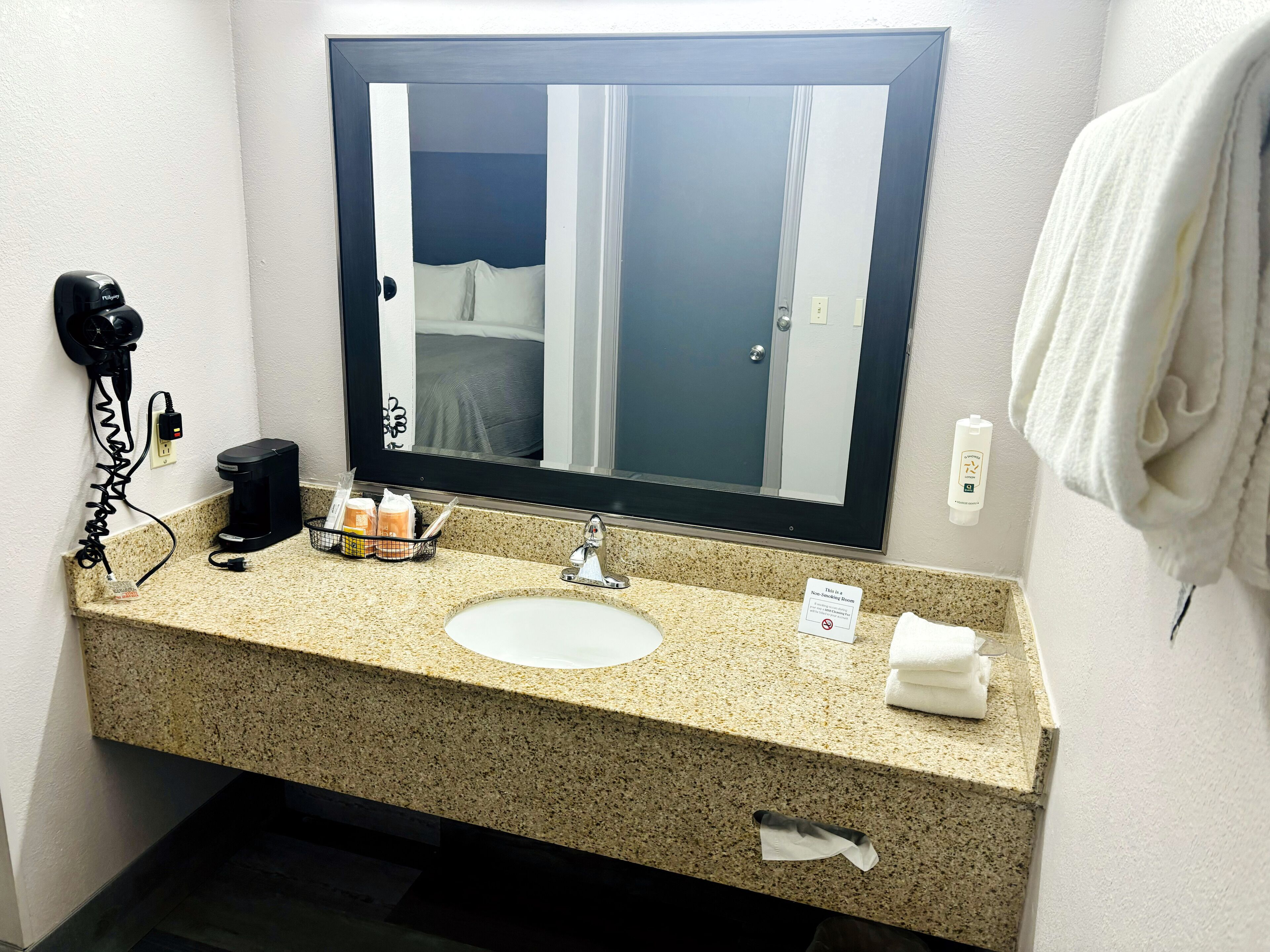 Casa de banho de hotel com lavatório de bancada em granito, um secador de cabelo preto, máquina de café e porta-toalhas.
