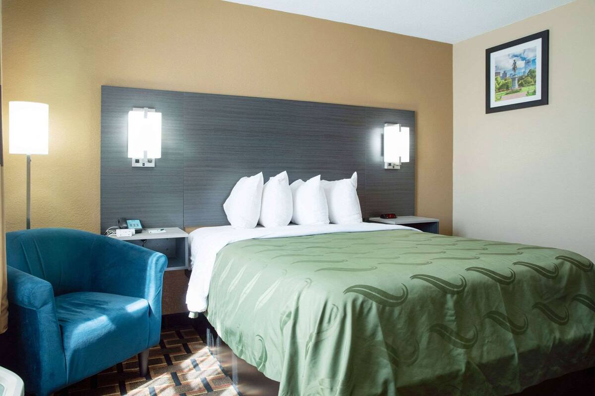 Quarto de hotel com cama, uma cadeira, mesa de cabeceira e luminária na parede.