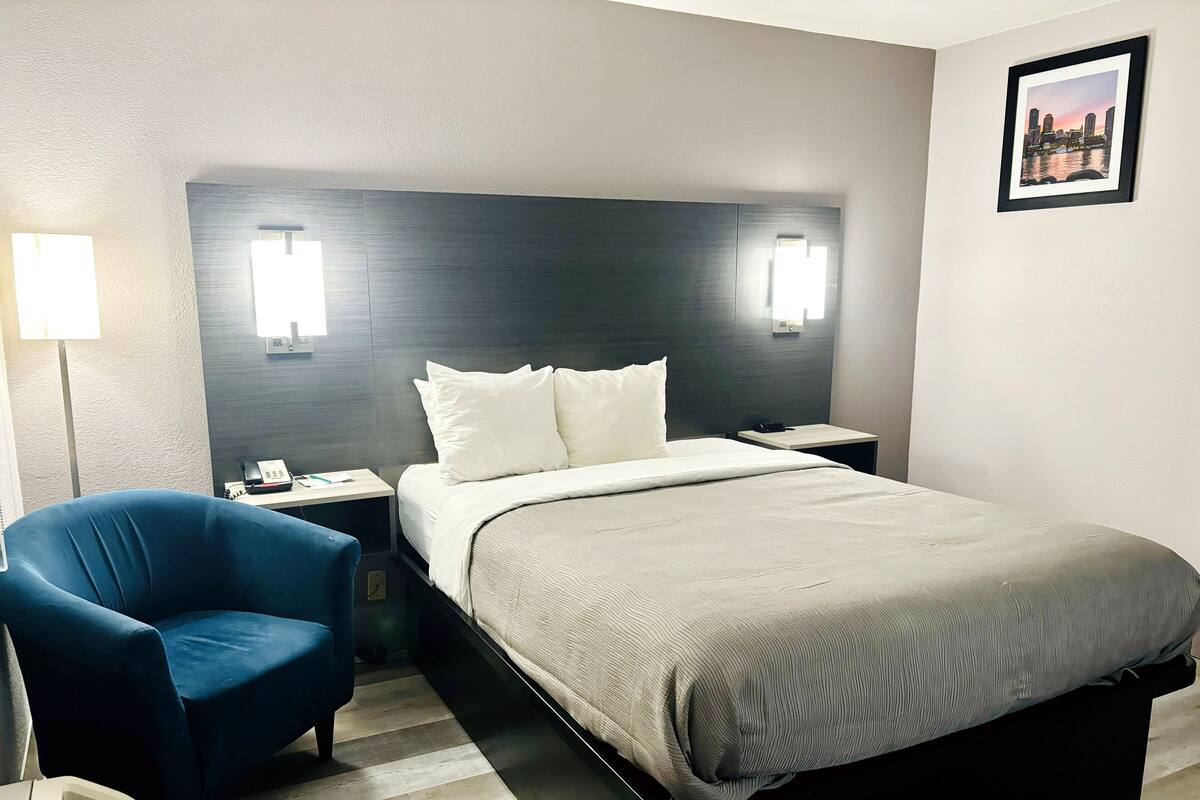Quarto de hotel com uma cama grande, uma poltrona azul, uma mesinha de cabeceira com telefone e um candeeiro fixado na parede.