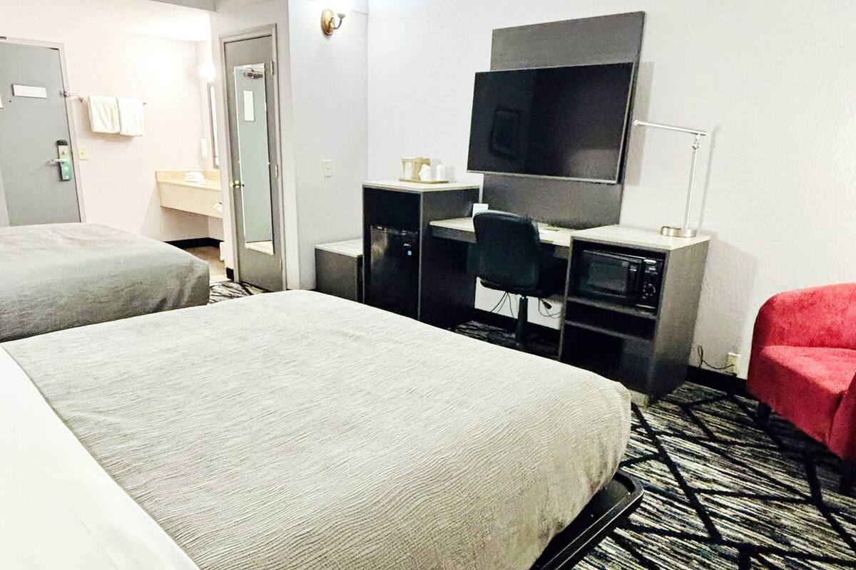 Quarto de hotel com duas camas, uma escrivaninha com computador, uma televisão e uma cadeira vermelha.