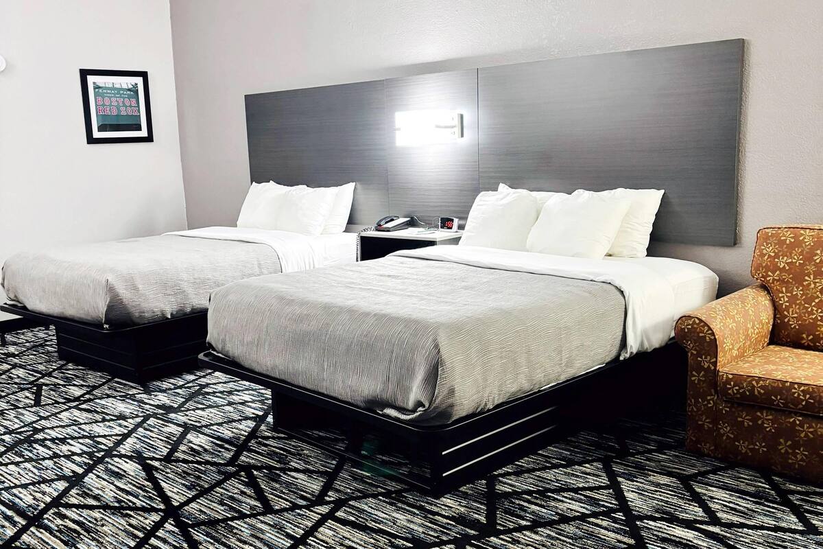Quarto de hotel com duas camas, uma mesa de cabeceira, uma cadeira e um tapete estampado.