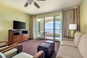 Quarto de hotel com televisão de tela plana em um móvel de madeira, sofá, um pequeno banquinho e varanda com vista para o mar.