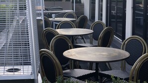 Uma área de estar externa com mesas e cadeiras redondas, uma estrutura de metal para churrasqueira e um edifício com uma placa que diz "MEO'S".