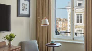 Quarto de hotel com cama, uma cadeira, uma mesinha com uma chávena e um livro, uma lâmpada, televisão e janela com vista para edifícios.