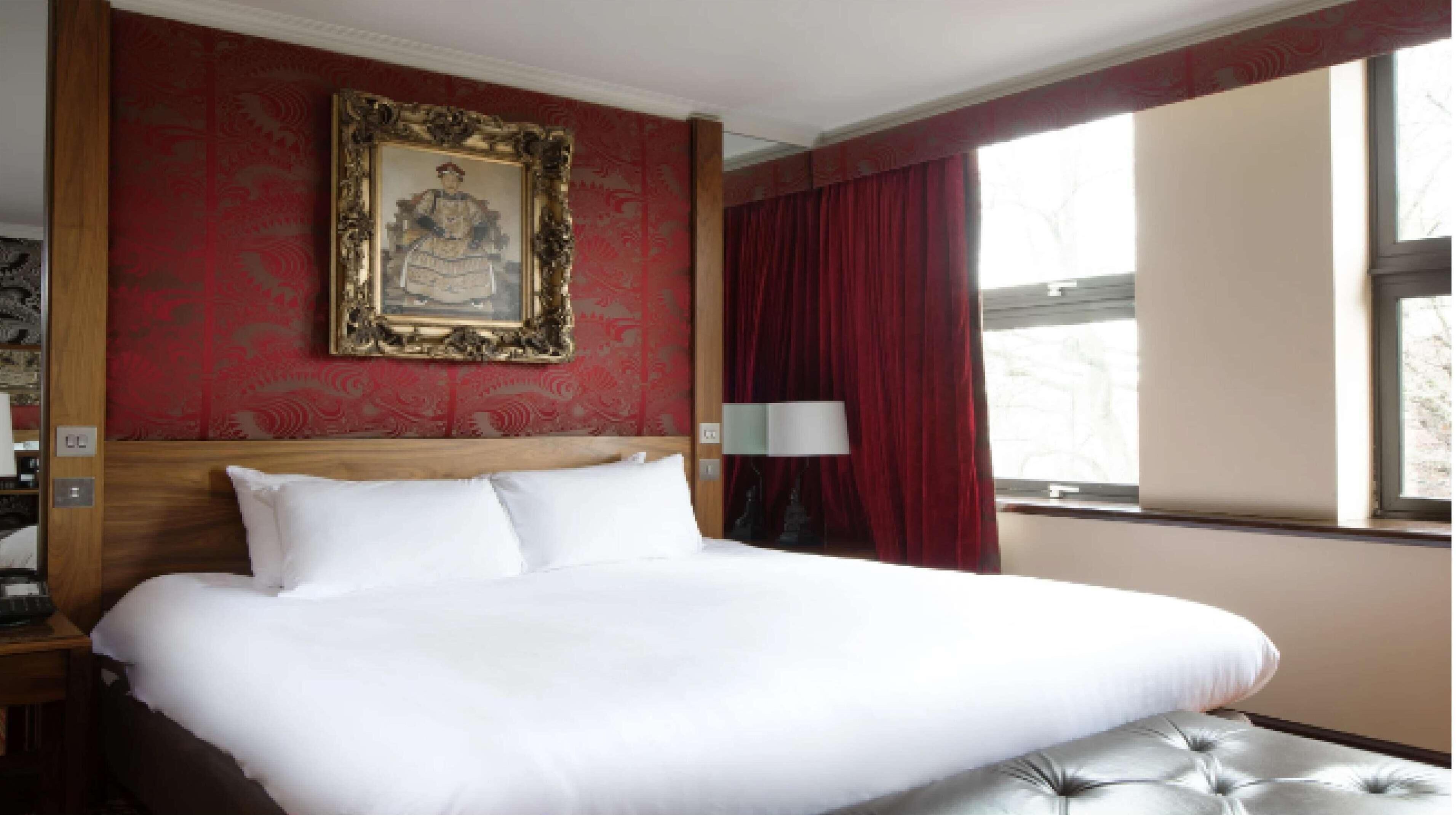 Quarto de hotel com uma cama grande, cabeceira de madeira, quadro na parede, cortina vermelha e janela com vista.