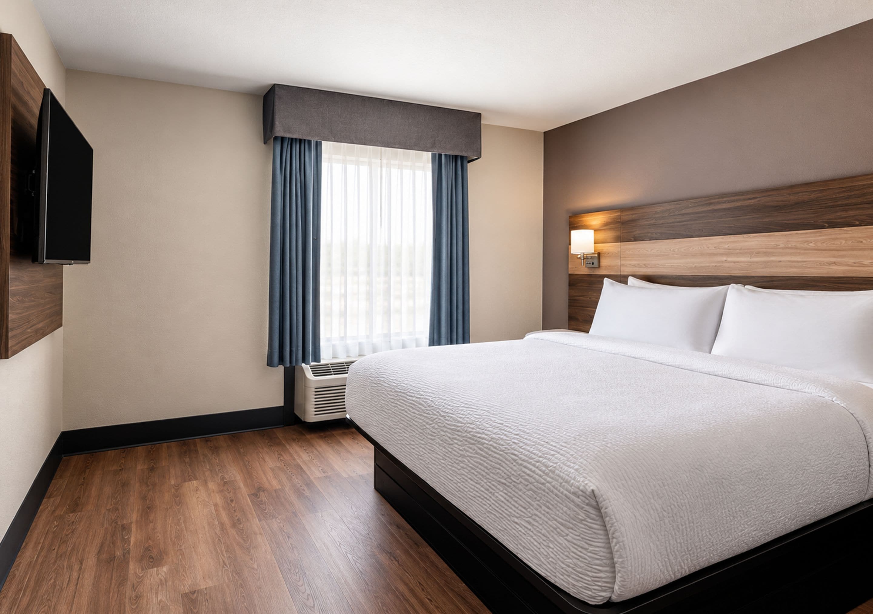 Quarto de hotel com cama, televisão de tela plana, janela com cortinas e luminária fixada na parede.