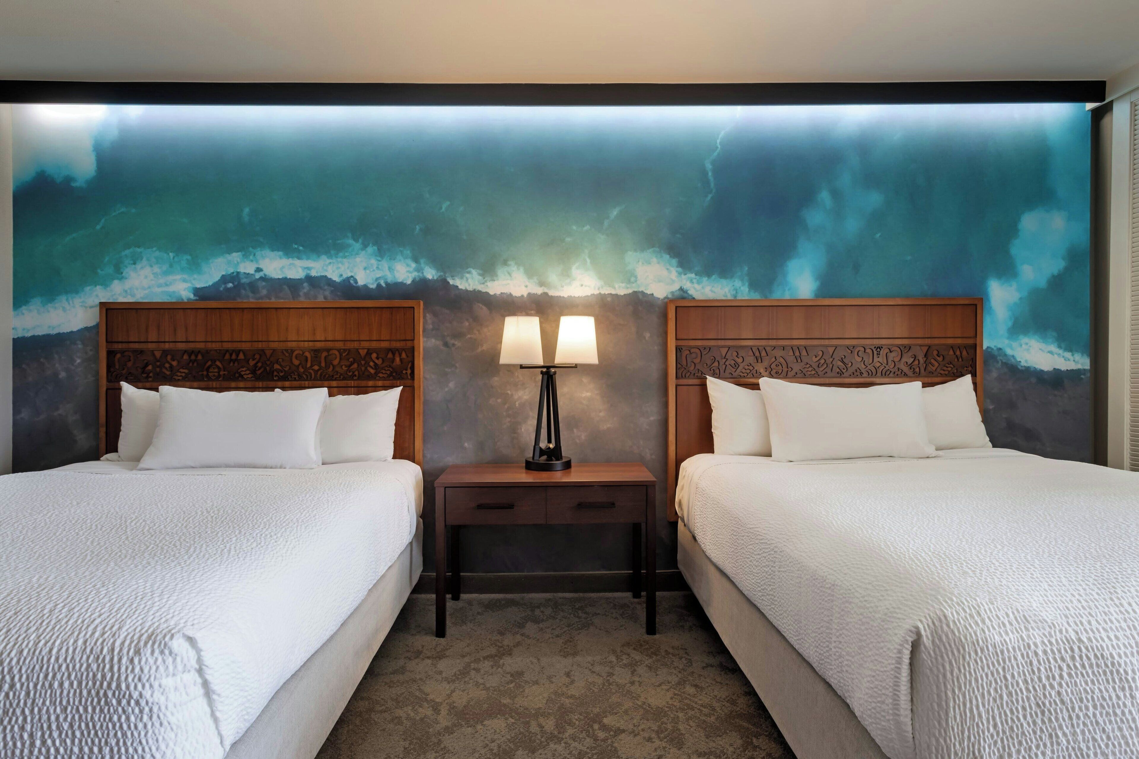 Quarto de hotel com duas camas, uma mesa de cabeceira com abajur e um grande mural na parede representando um mar tempestuoso.