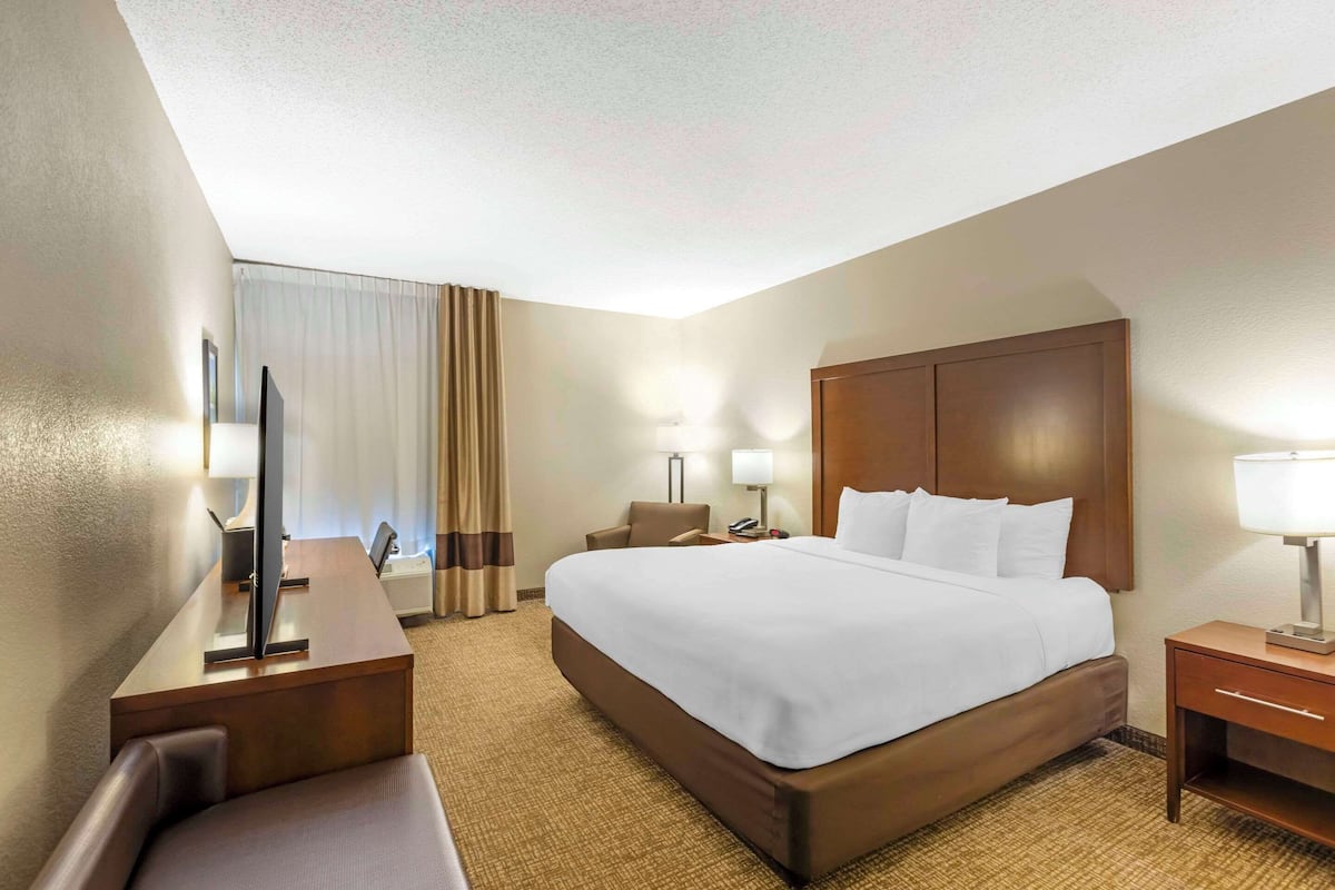 Quarto de hotel com uma cama grande, uma escrivaninha com computador, uma cadeira e uma mesa de cabeceira com abajur.