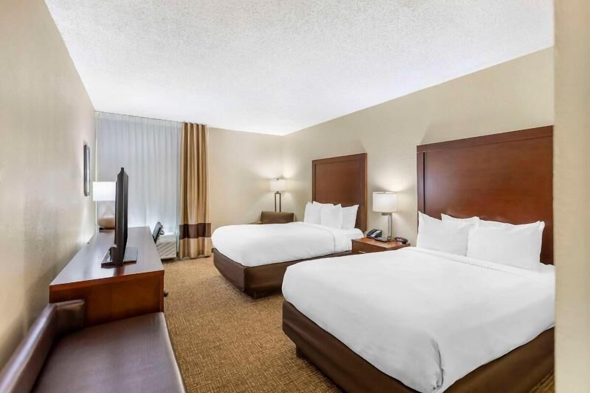 Quarto de hotel com duas camas, uma escrivaninha, televisão e janela com cortinas.