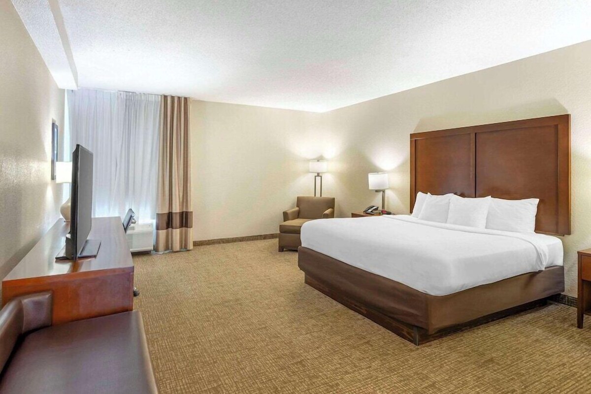 Quarto de hotel com uma cama grande, uma escrivaninha, uma cadeira, uma televisão e um abajur.