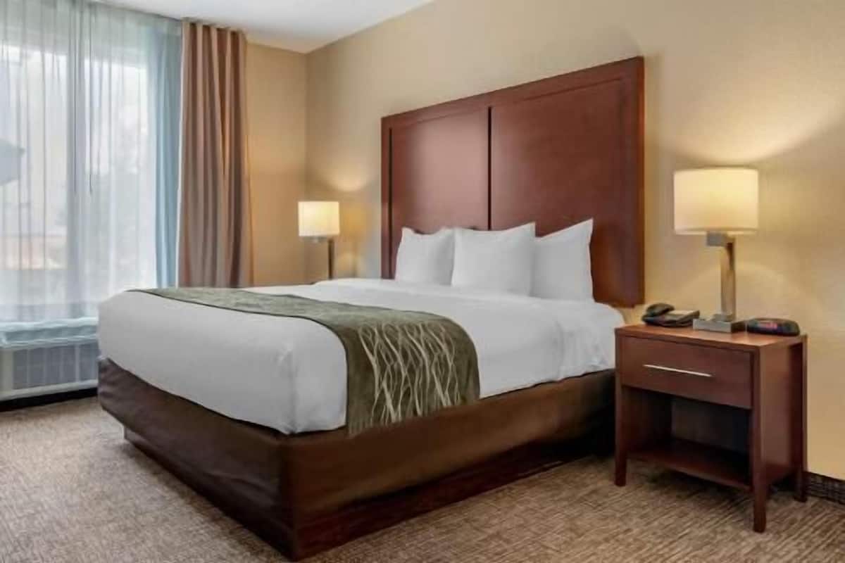 Quarto de hotel com uma cama grande, duas luminárias de cabeceira, uma mesinha de madeira e uma janela com cortinas.