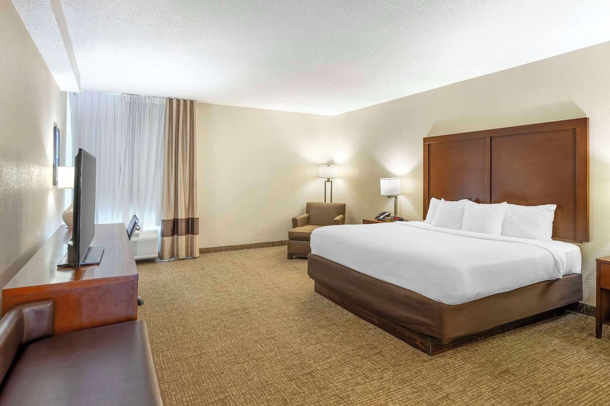 Quarto de hotel com uma cama grande, uma escrivaninha com televisão de tela plana, uma cadeira, um abajur e uma mesa de cabeceira.