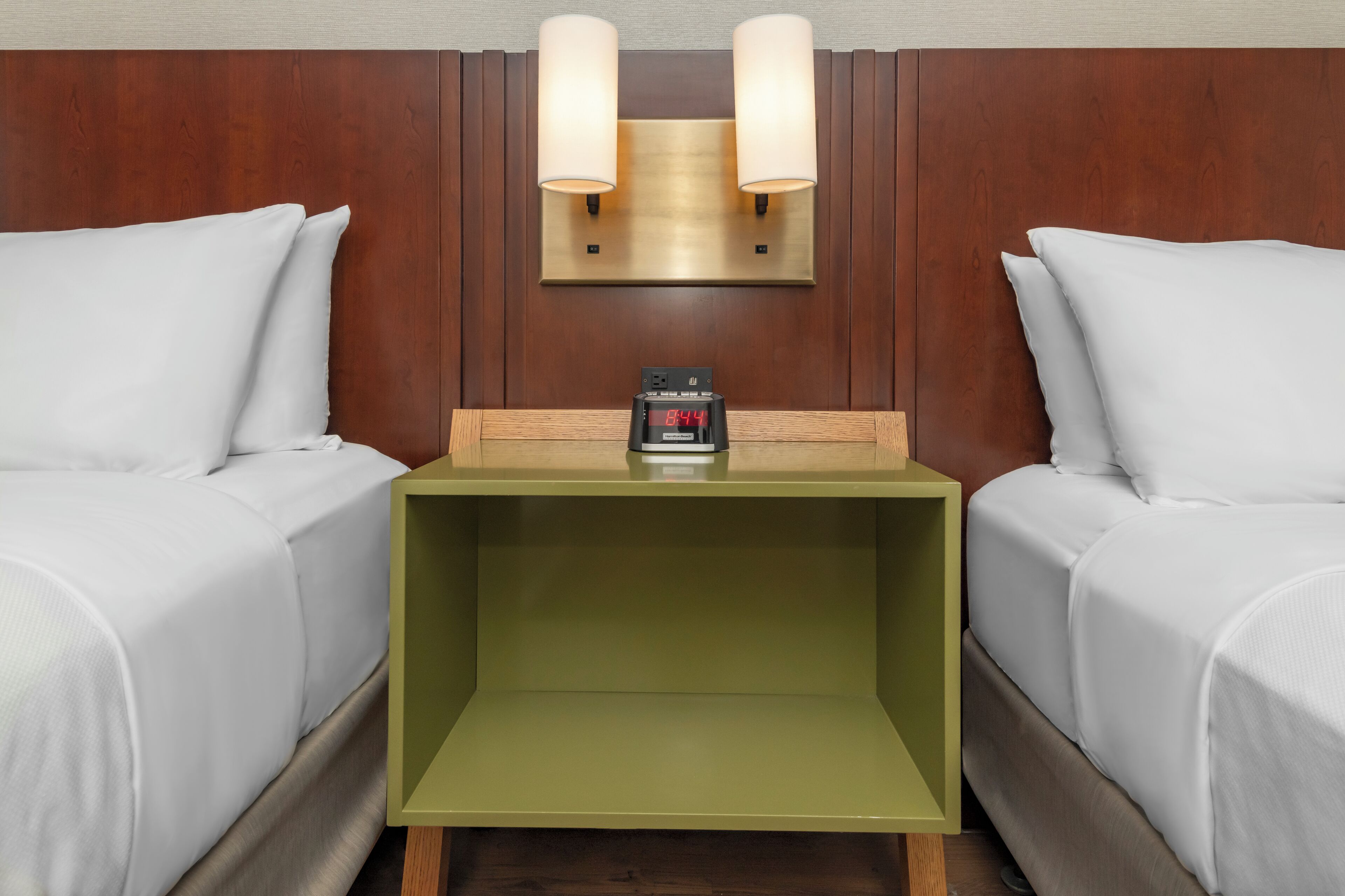 Quarto de hotel com duas camas, uma mesa de cabeceira verde, um relógio digital e luminárias fixadas na parede.