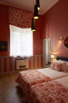 Quarto com duas camas, janela com cortinas, radiador, guarda-roupa, mesa de cabeceira com abajur e espelho na parede.