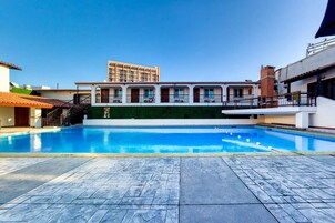 Uma piscina com deck revestido, um edifício com várias varandas e um prédio de apartamentos alto ao fundo.