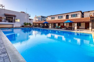 Piscina com espreguiçadeiras e guarda-sóis, cercada por edifícios com varandas e janelas.