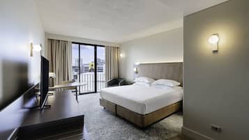 Quarto de hotel com cama, escrivaninha, cadeira, televisão e varanda com vista para a cidade.
