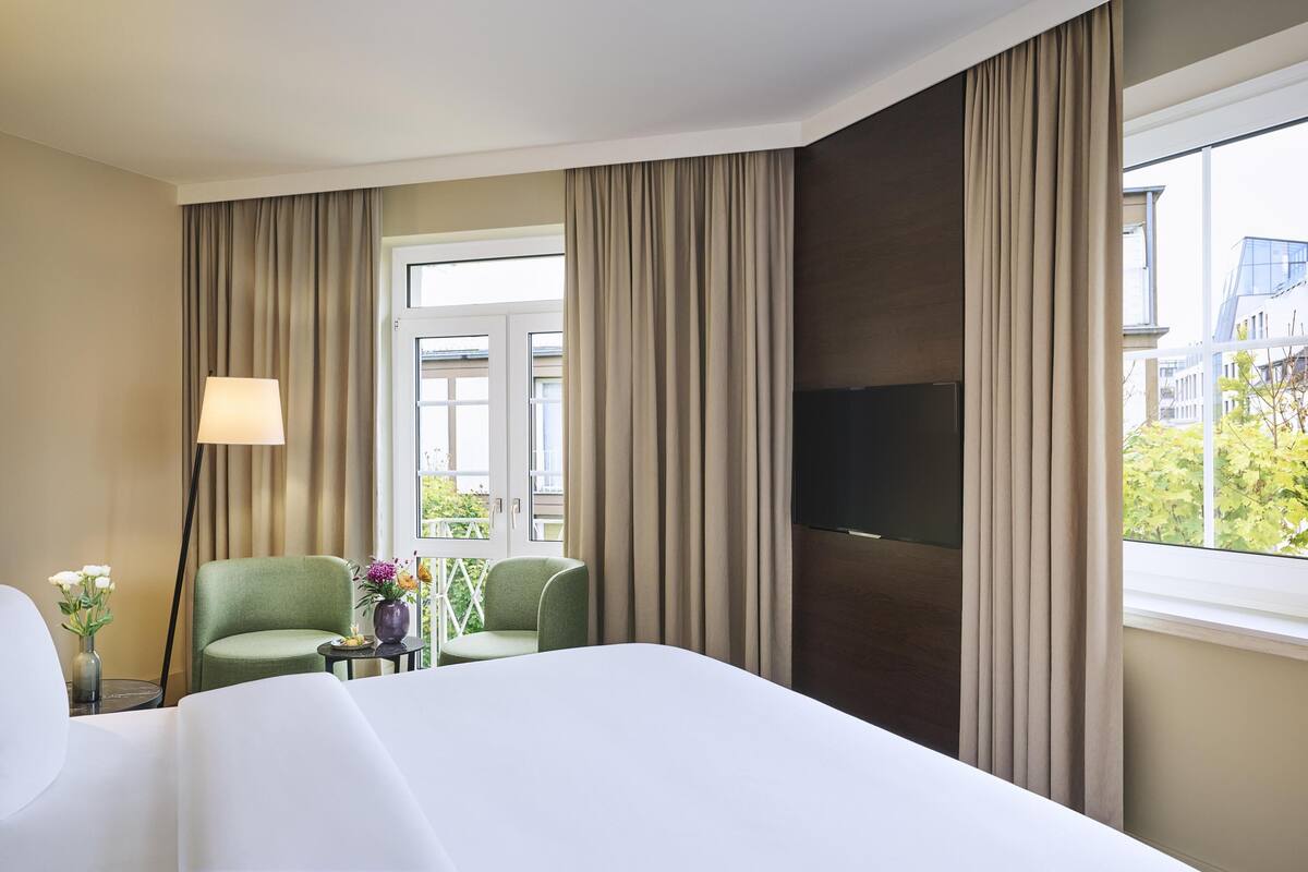 Quarto de hotel com cama, duas cadeiras, televisão e janela com cortinas.