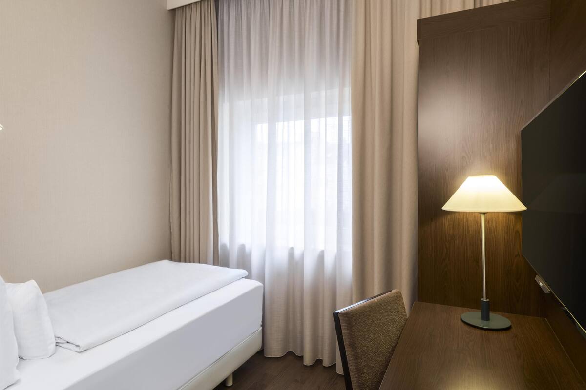 Quarto de hotel com cama, poltrona, escrivaninha, abajur e televisão.