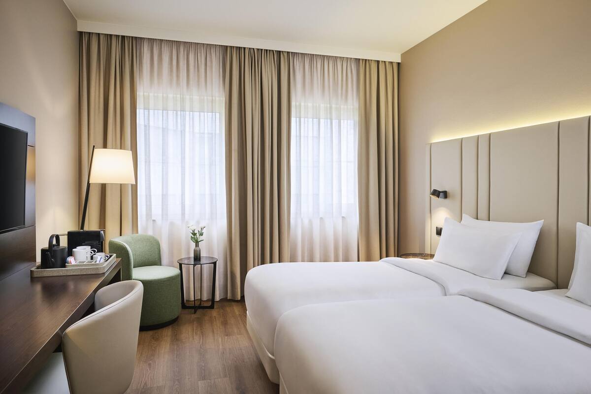 Quarto de hotel com duas camas, uma escrivaninha, uma cadeira, um abajur e uma pequena mesa com uma planta.