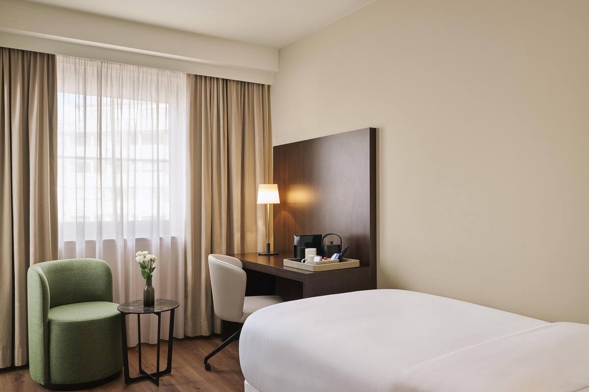 Quarto de hotel com uma cama, uma escrivaninha com abajur, uma cadeira e uma mesa pequena com um vaso de flores.
