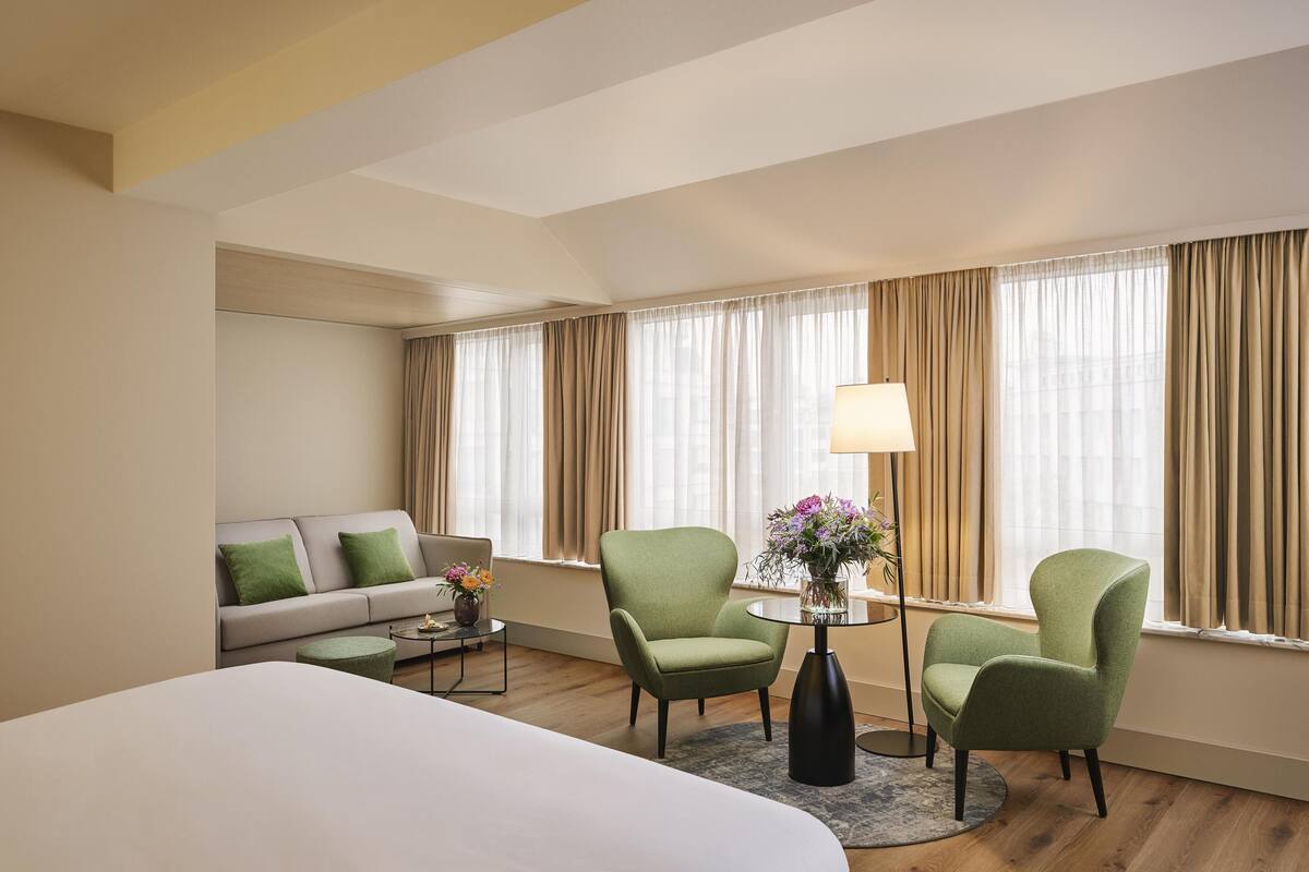 Quarto de hotel com uma cama, um sofá, duas poltronas, uma mesinha de centro com abajur e um vaso com flores.