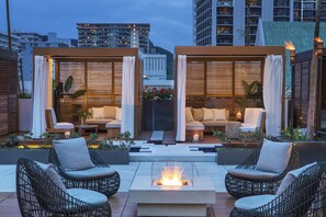 Terraço no telhado com móveis de vime, uma lareira central e vista para prédios da cidade.