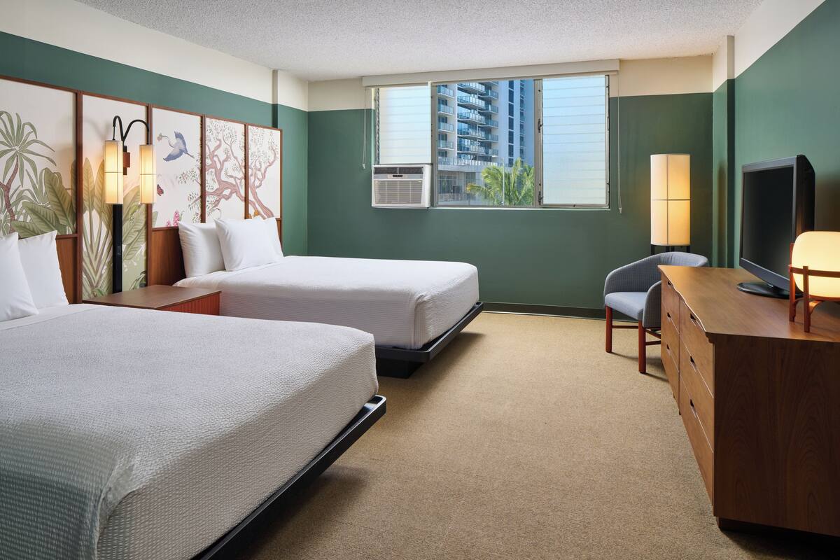 Quarto de hotel com duas camas, uma escrivaninha, uma cadeira, uma televisão e janela com vista para edifícios.