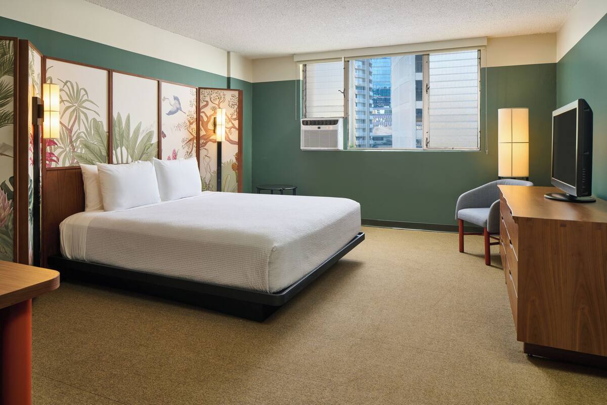 Quarto de hotel com uma cama, escrivaninha, cadeira, televisão e janela com persianas.