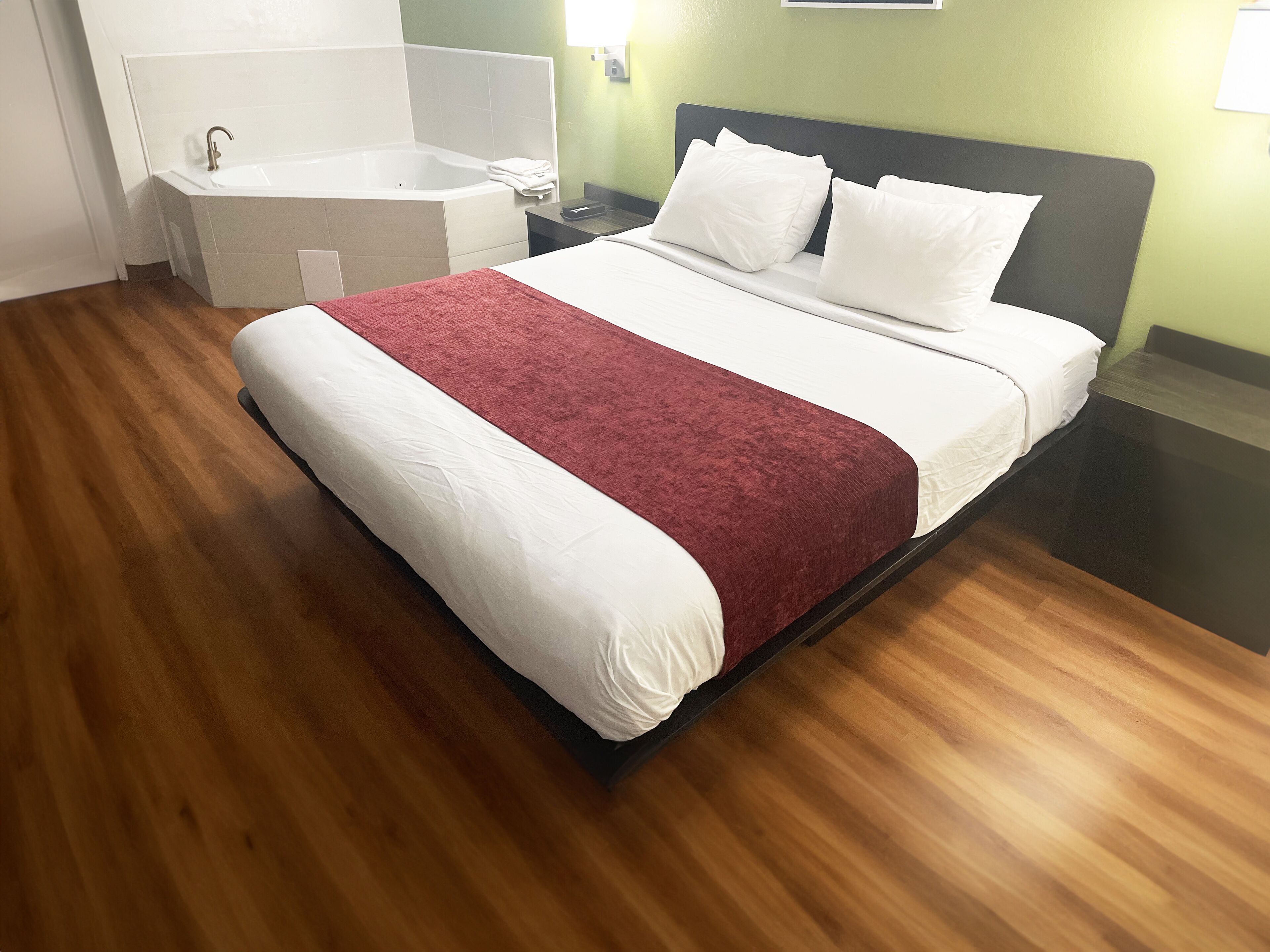 Quarto de hotel com cama, manta vermelha, travesseiro branco, mesa de cabeceira e banheiro com banheira.
