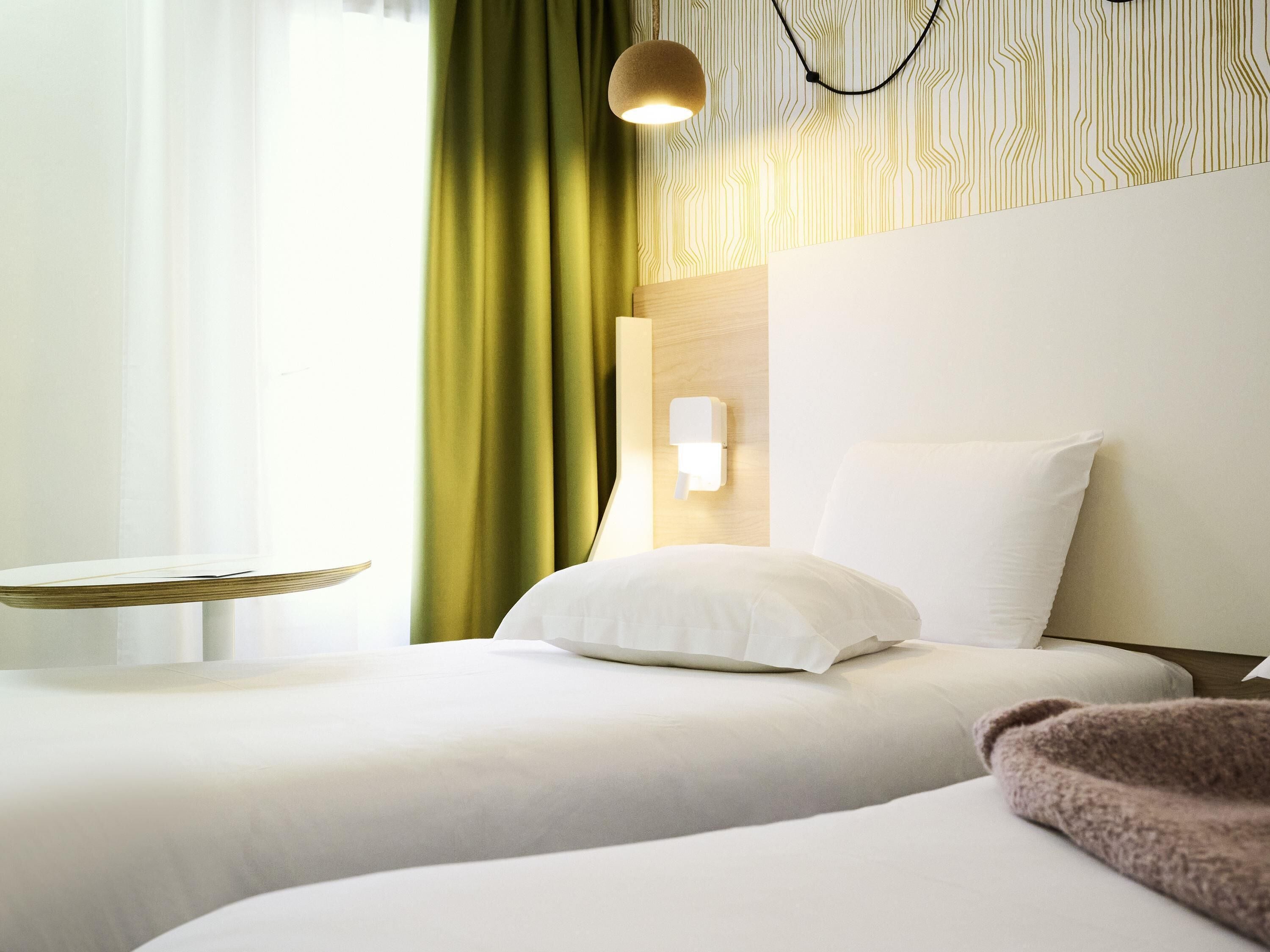 Quarto de hotel com duas camas, uma mesa redonda, uma luminária na parede e um papel de parede estampado.