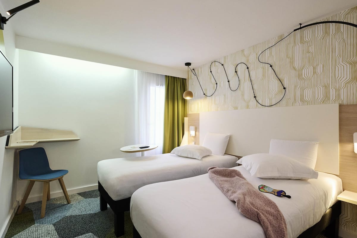 Quarto de hotel com duas camas, uma escrivaninha, uma cadeira e um papel de parede estampado.