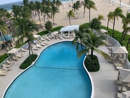 Piscina com espreguiçadeiras e guarda-sóis, cercada por palmeiras e vista para a praia.