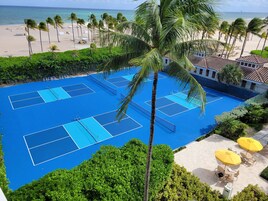 Uma quadra de tênis azul com piscina, cercada por palmeiras e cadeiras de praia.