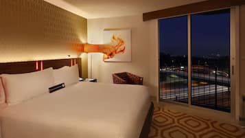 Quarto de hotel com uma cama grande, mesa de cabeceira, luminária, cadeira e uma janela ampla com vista para a cidade.