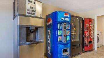 Uma área com máquinas de venda automática, incluindo uma máquina de Pepsi, uma de Coca-Cola e um bebedouro.