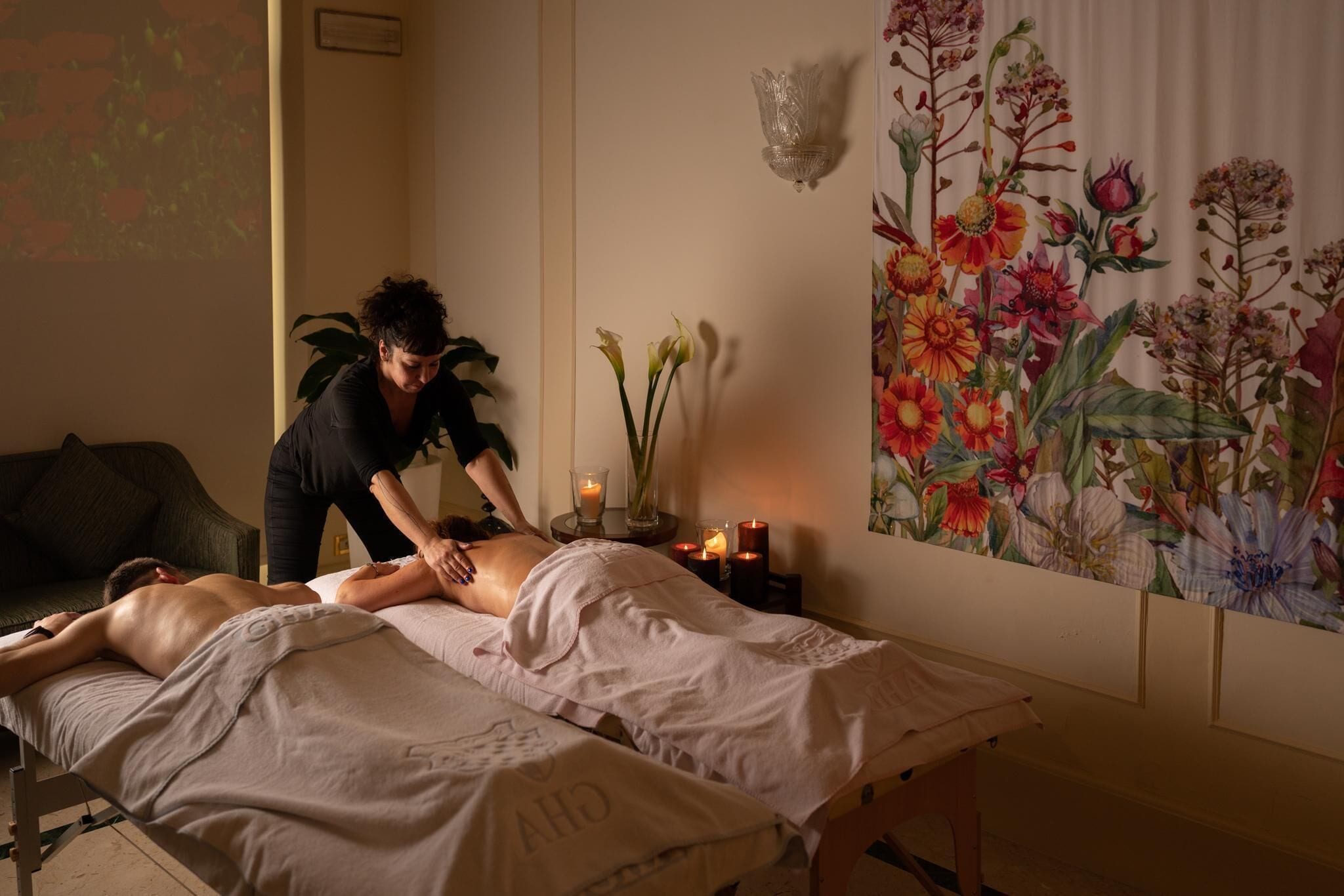 Sala de massagem com mesa de massagem, um cliente deitado de bruços, terapeuta atendendo o cliente, parede com tapeçaria floral e uma mesinha com velas.