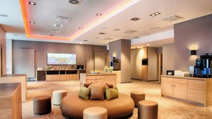 Um lobby de hotel moderno com área de estar central, mobiliário de madeira e uma grande TV de tela plana na parede.