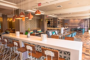 Uma área de bar moderna com um balcão comprido, diversas opções de assento, incluindo banquetas e mesas, e iluminação pendente.