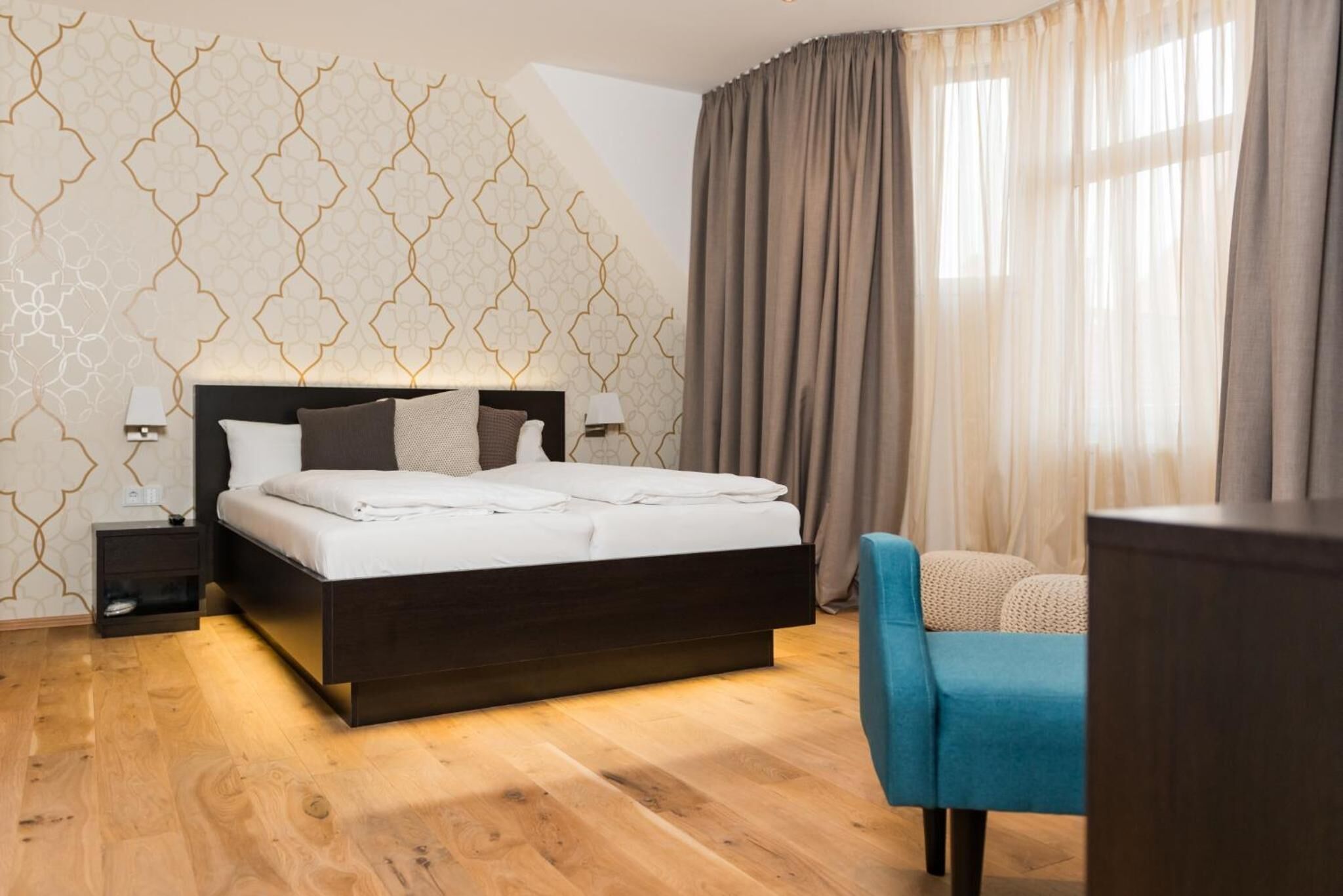 Quarto moderno com cama grande, mesas de cabeceira, uma cadeira azul e papel de parede estampado.