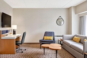 Quarto de hotel com escrivaninha, cadeira, sofá, duas mesinhas, televisão e espelho.