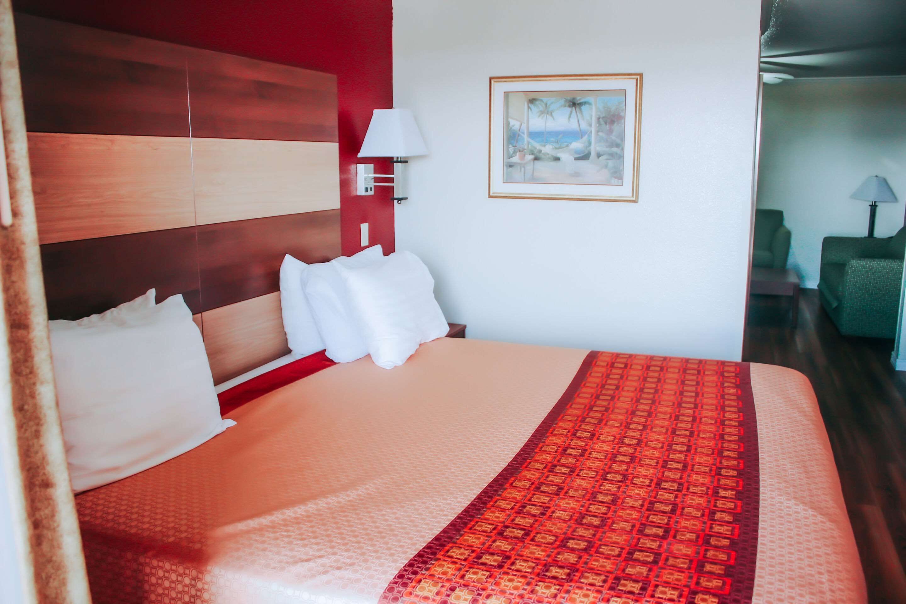 Quarto de hotel com uma cama grande, cabeceira de madeira, parede de destaque vermelha, quadro pendurado na parede e poltrona verde no canto.