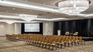 Uma sala de conferências com fileiras de cadeiras, palco e uma tela grande exibindo "SHERATON".