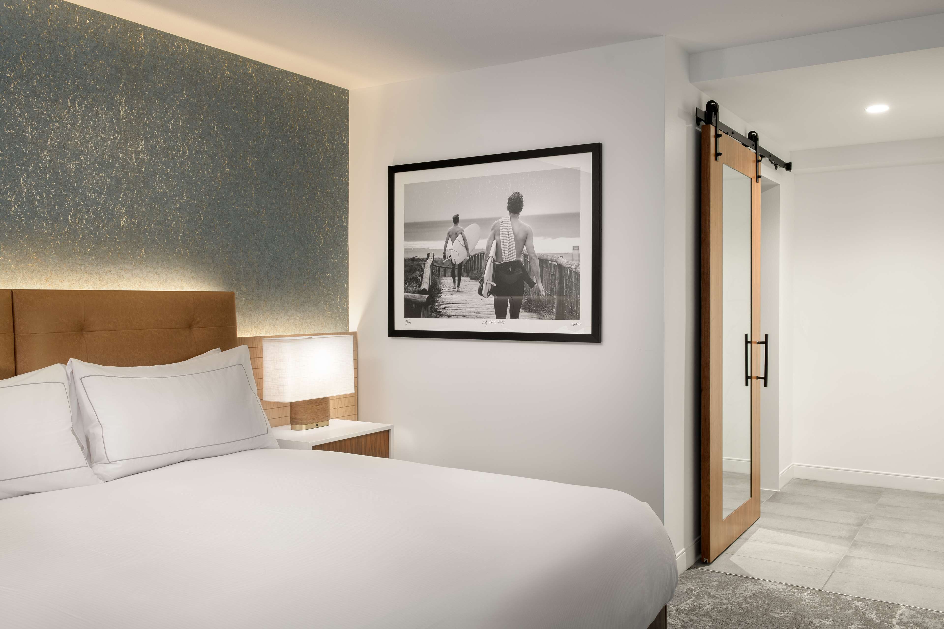 Quarto de hotel com uma cama grande, um quadro pendurado na parede, uma porta de madeira e uma mesa de cabeceira com abajur.