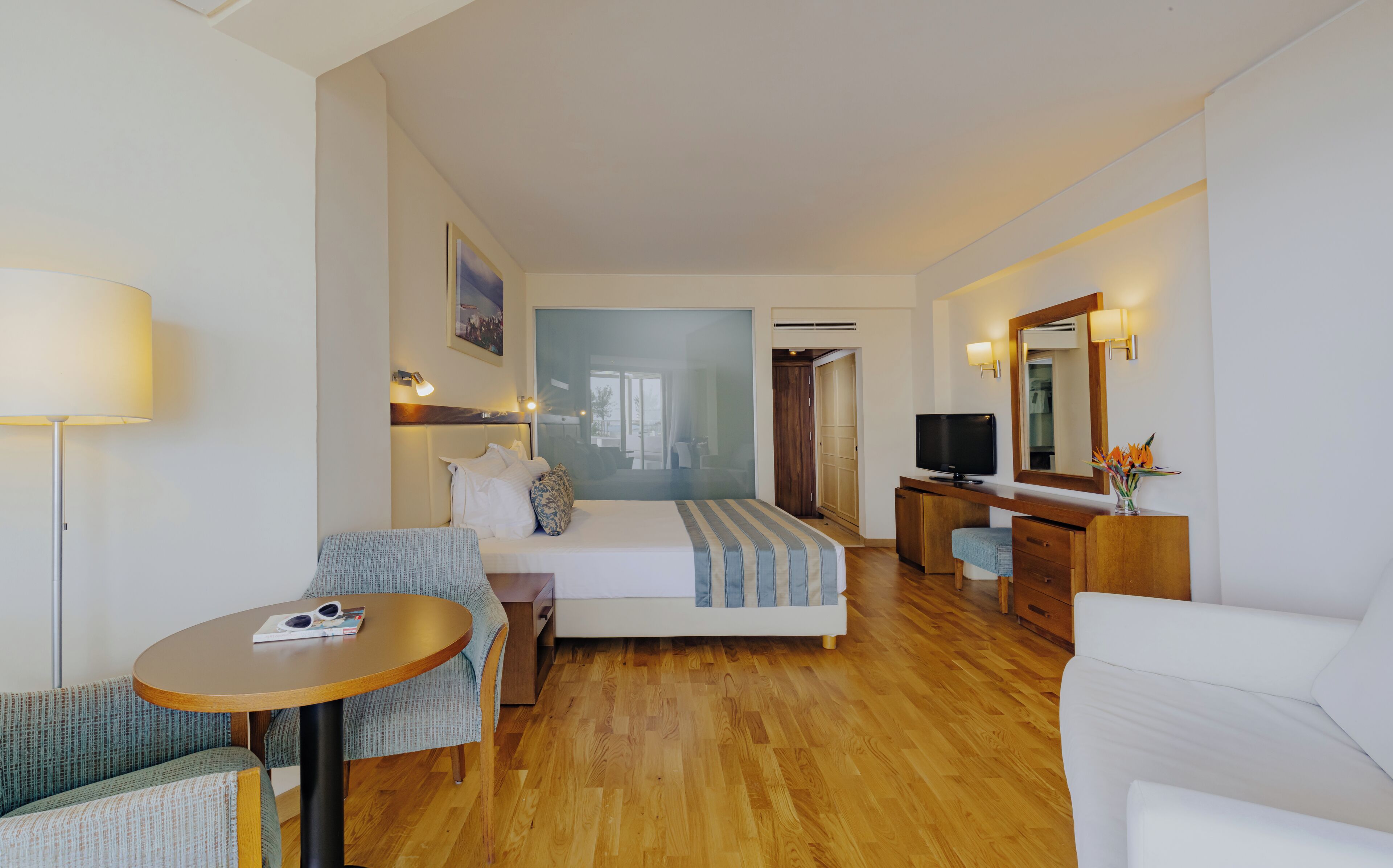 Quarto de hotel com cama, escrivaninha, cadeira, mesinha e televisão.