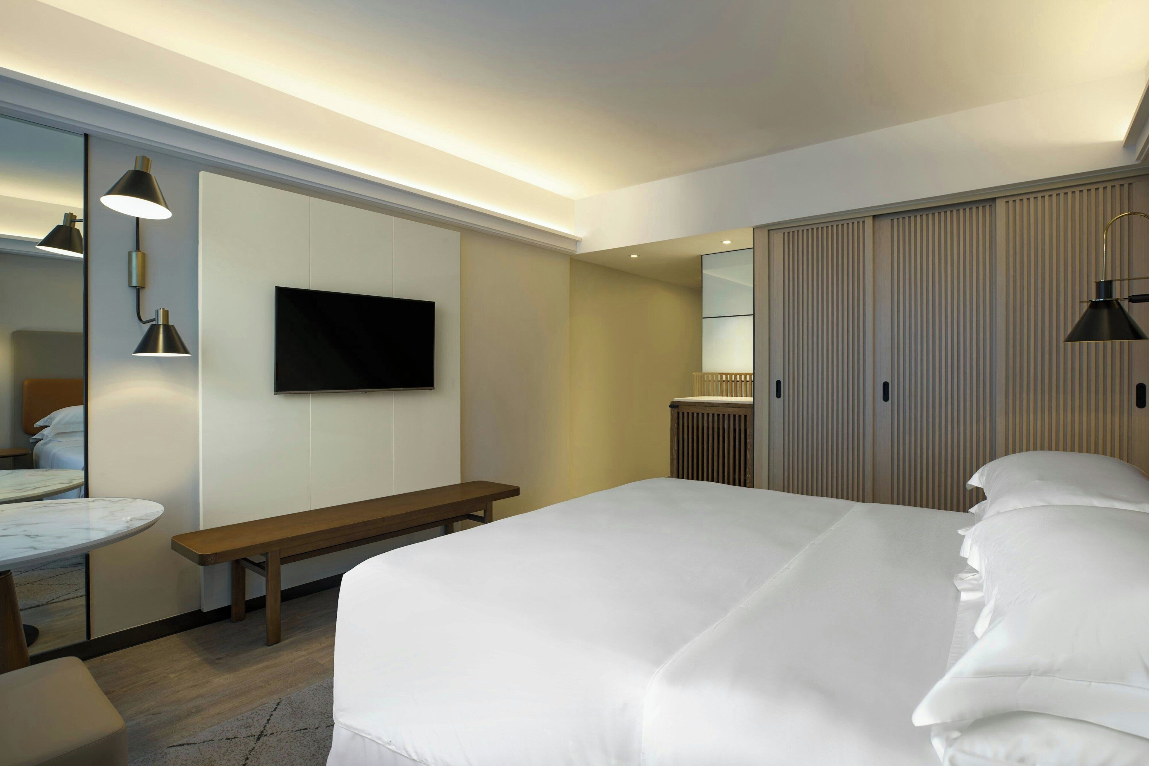 Quarto de hotel com uma cama grande, mesa de cabeceira, espelho, televisão na parede e um banco.