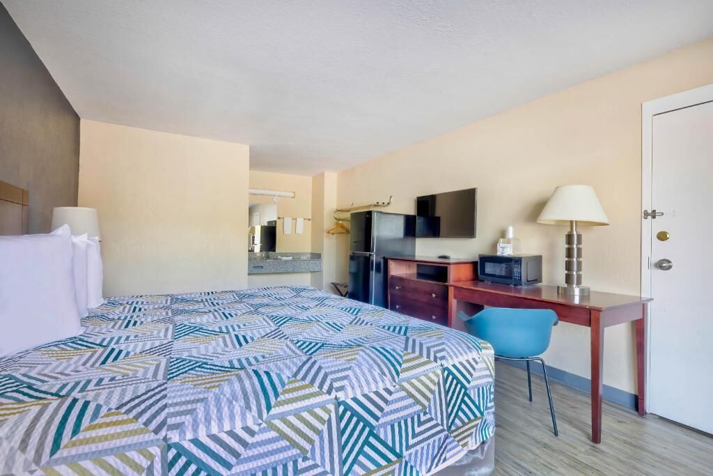 Quarto de hotel com cama, escrivaninha com cadeira, televisão, micro-ondas e porta.