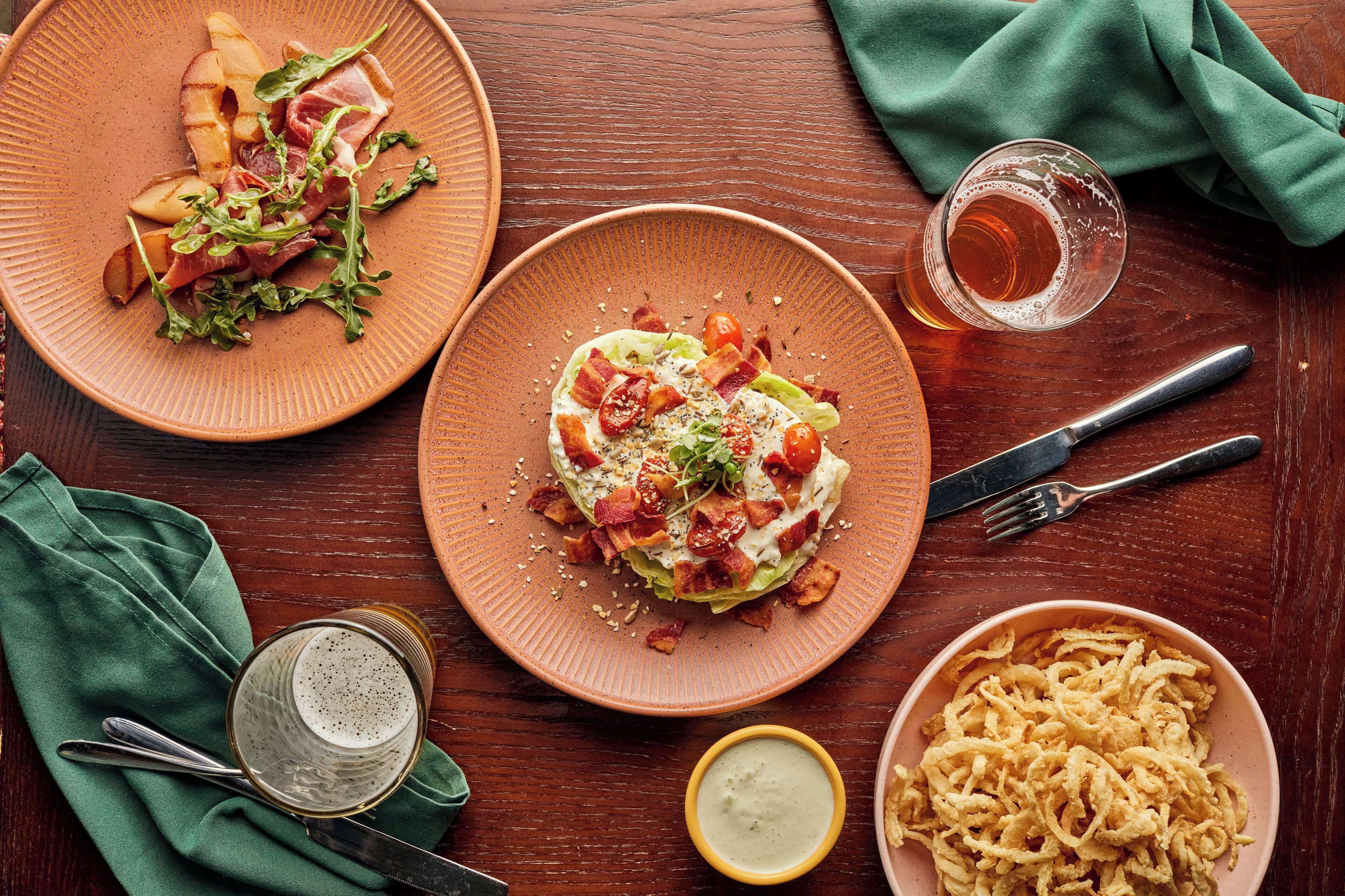 Uma mesa de madeira com pratos de comida, um copo de cerveja e talheres.