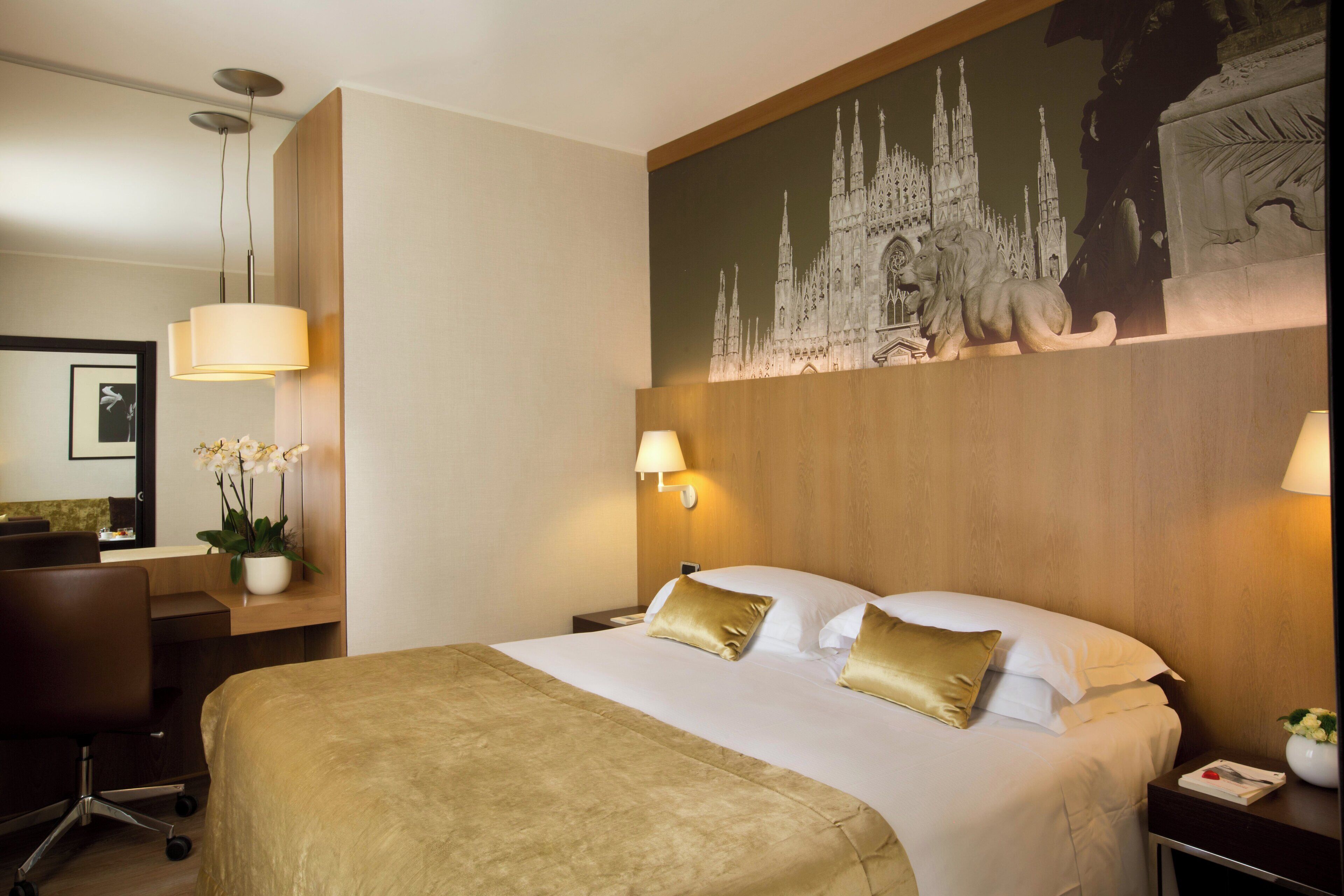 Quarto de hotel com uma cama grande, duas luminárias de cabeceira, uma escrivaninha com cadeira e um quadro na parede representando um castelo.